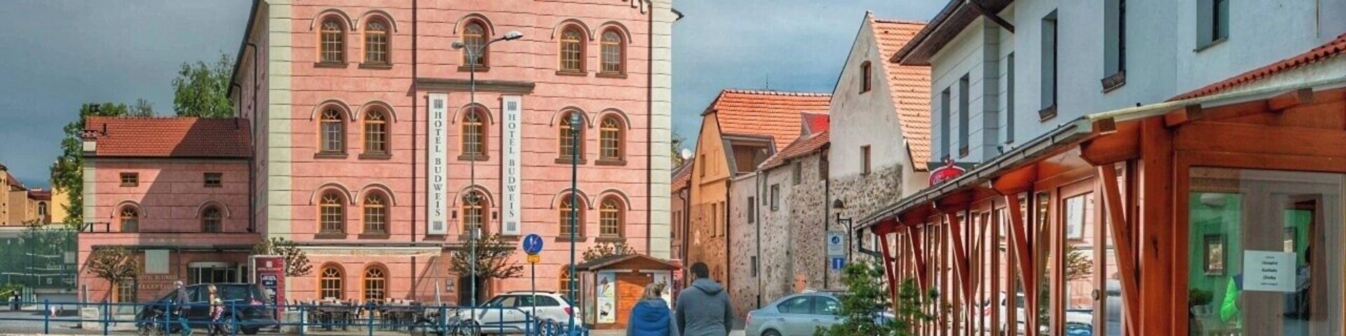 Um edifício histórico com janelas em arco e fachada vermelha, um canal com guarda-corpo azul e um café com toldo vermelho.