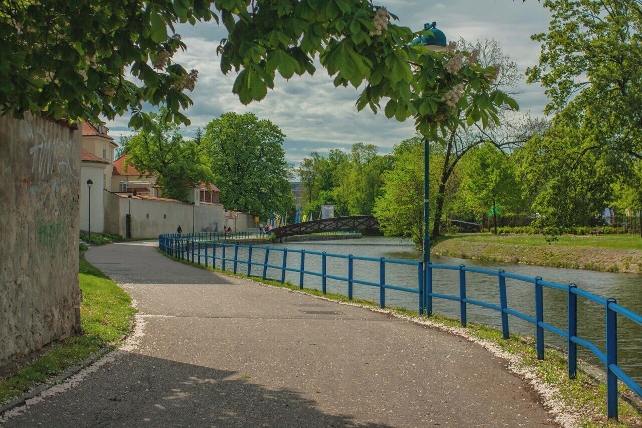 Um caminho pavimentado ao longo de um rio, com guarda-corpo azul, uma ponte e árvores ao fundo.