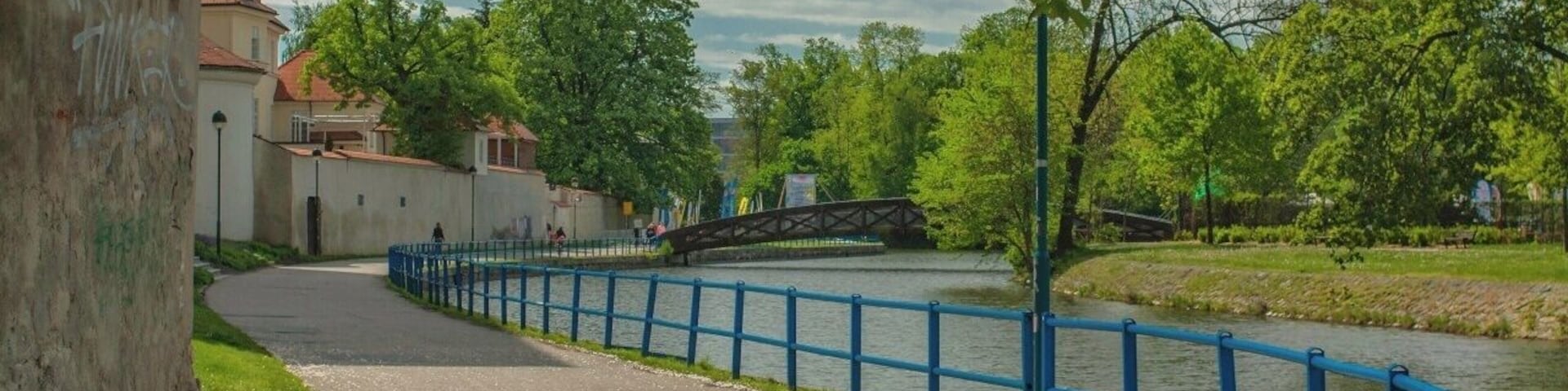 Um caminho pavimentado ao longo de um rio, com guarda-corpo azul, uma ponte e árvores ao fundo.