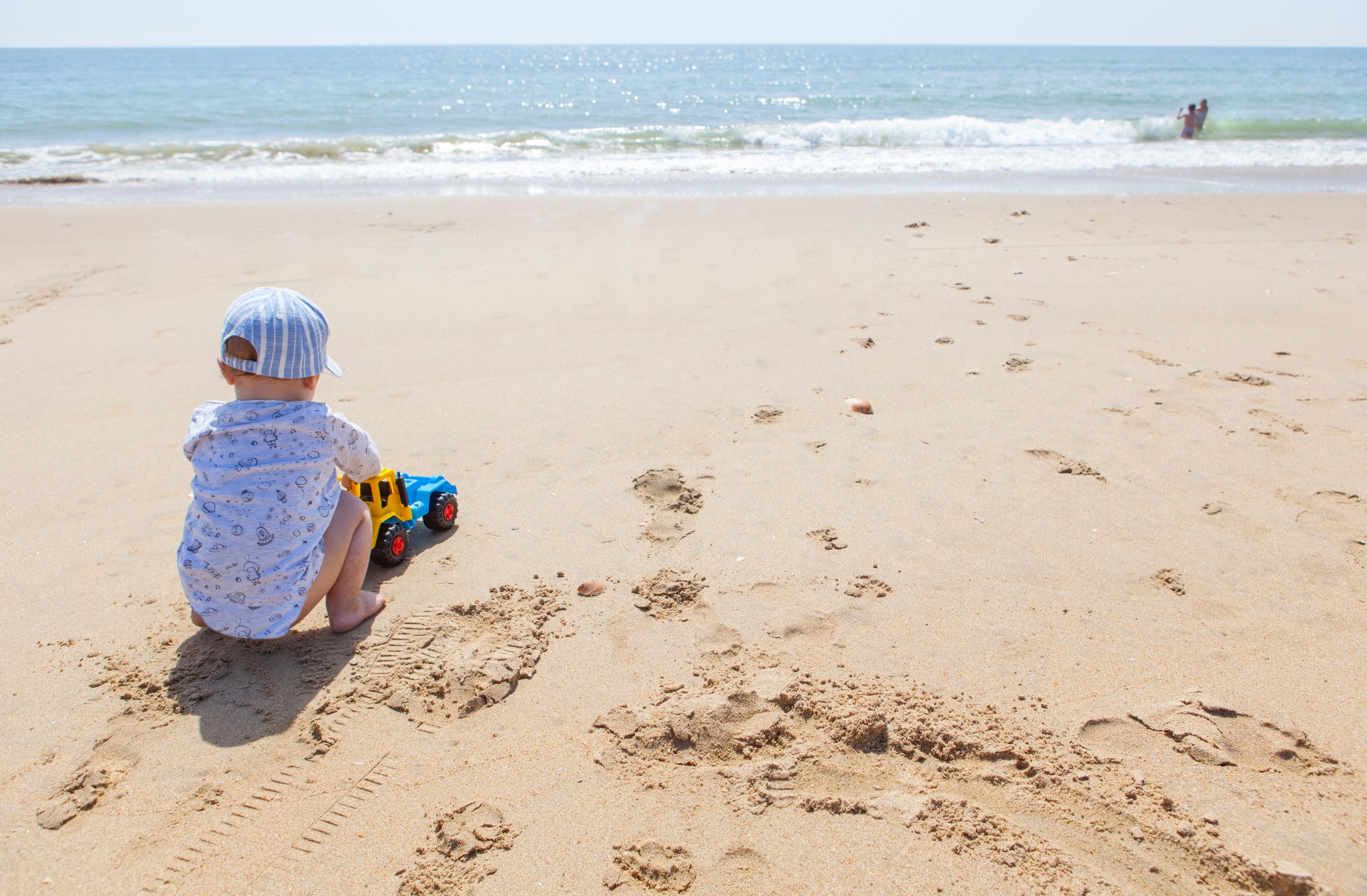 Criança brincando com um carrinho de brinquedo em uma praia arenosa, com pegadas e o oceano ao fundo.