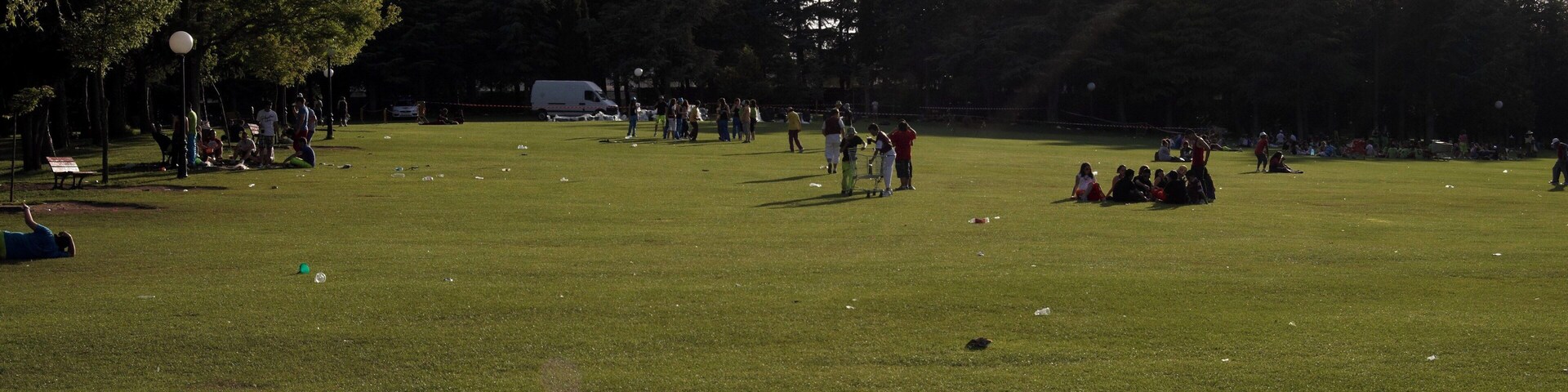 Um parque gramado com árvores, pessoas caminhando e alguns objetos espalhados pelo chão.
