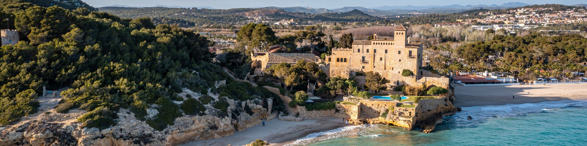 Uma vila costeira com um castelo imponente numa colina, águas turquesa cristalinas e uma praia de areia.