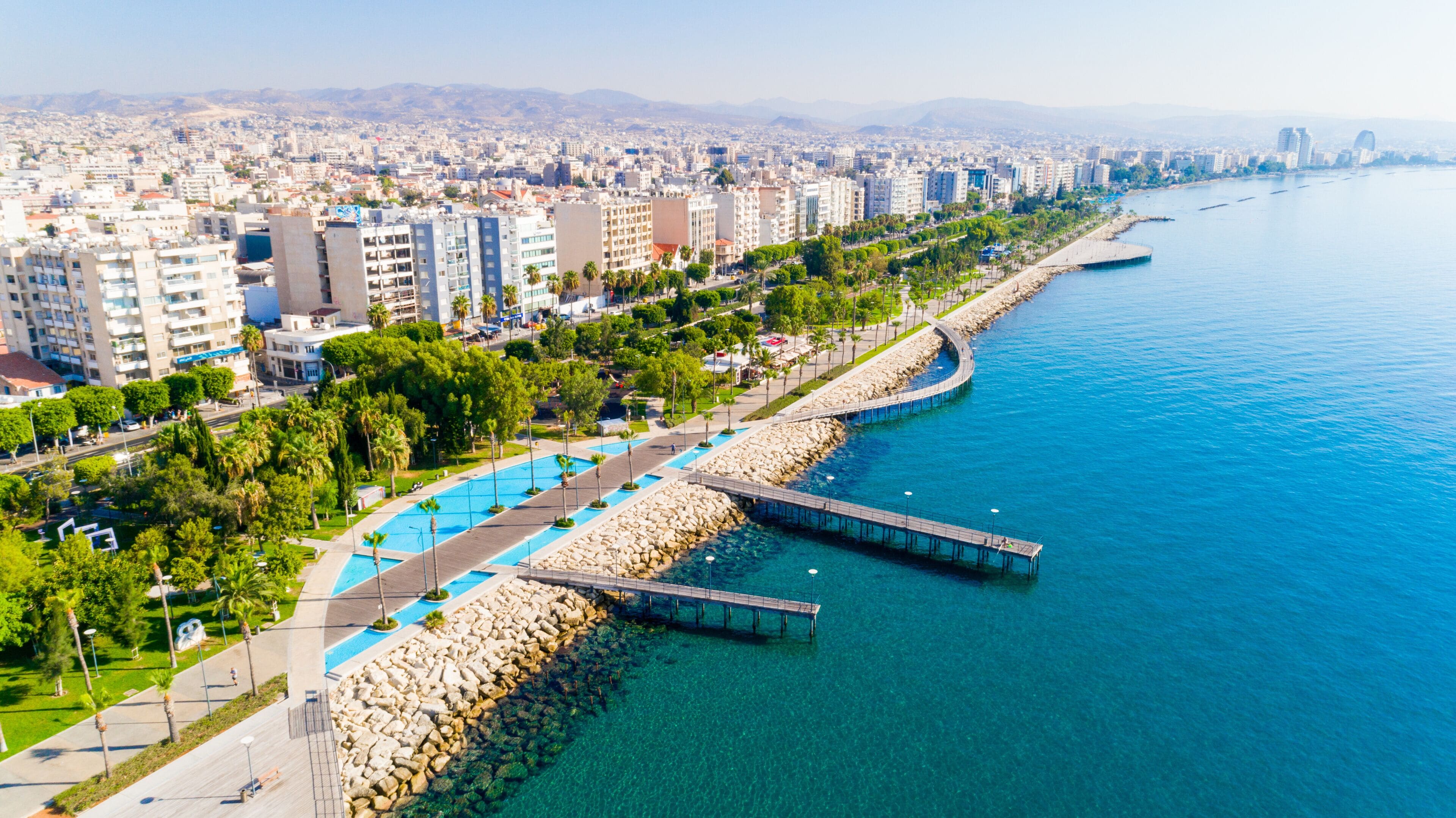 Paisagem costeira urbana com uma ampla orla marítima, mar azul límpido e uma cadeia montanhosa ao longe.