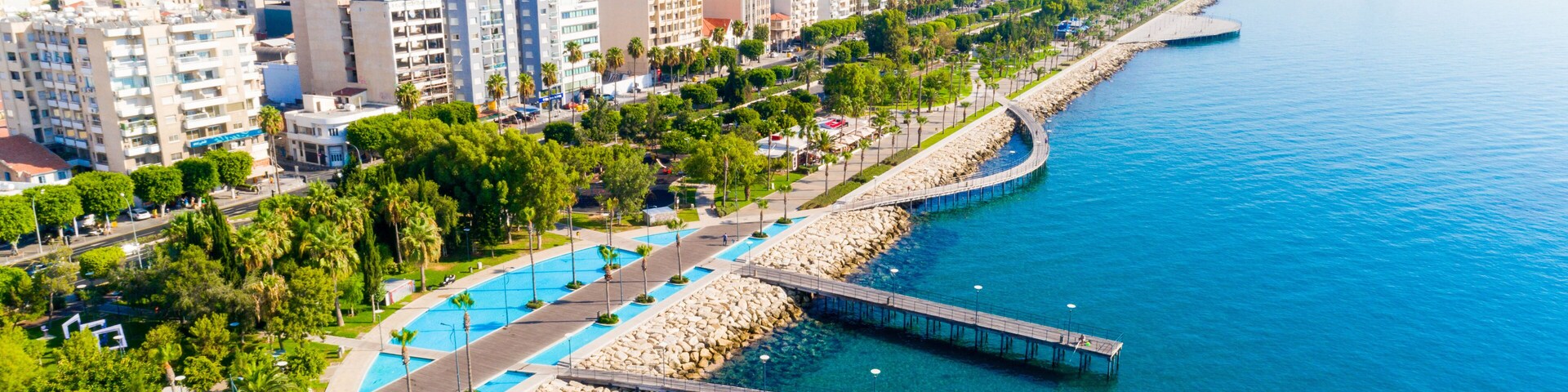Paisagem costeira urbana com uma ampla orla marítima, mar azul límpido e uma cadeia montanhosa ao longe.
