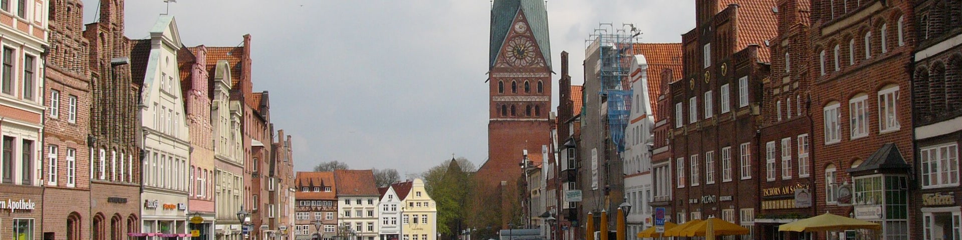 Ruas de paralelepípedos alinhadas por edifícios históricos, com uma torre de igreja imponente e alguns carros estacionados.