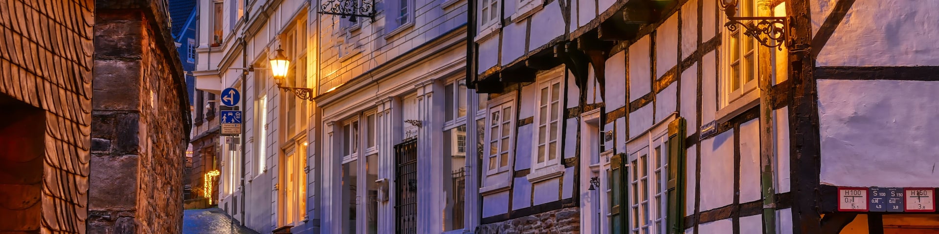 Uma rua de paralelepípedos com edifícios tradicionais em estilo enxaimel, uma placa de rua e um lampião.