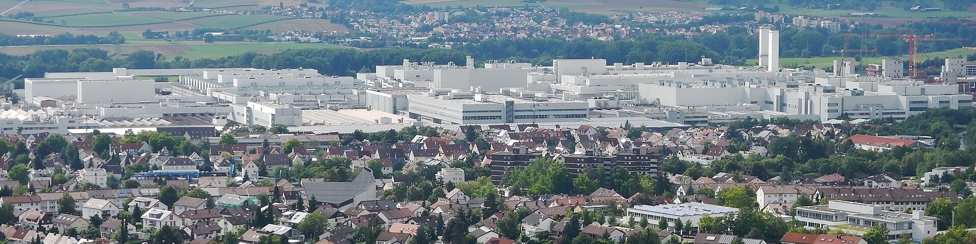 Paisagem urbana com mistura de edifícios residenciais e industriais, rodeada por campos verdes e colinas ao longe.