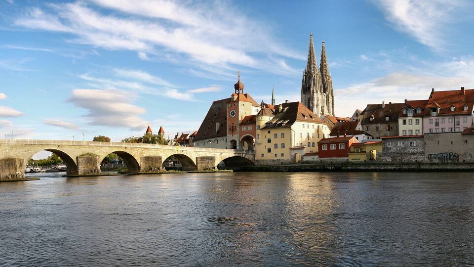 Uma ponte histórica com arcos, uma catedral com duas torres e edifícios coloridos ao longo de um rio.