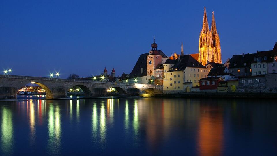 Uma ponte histórica, uma catedral e um rio refletindo luzes da cidade.