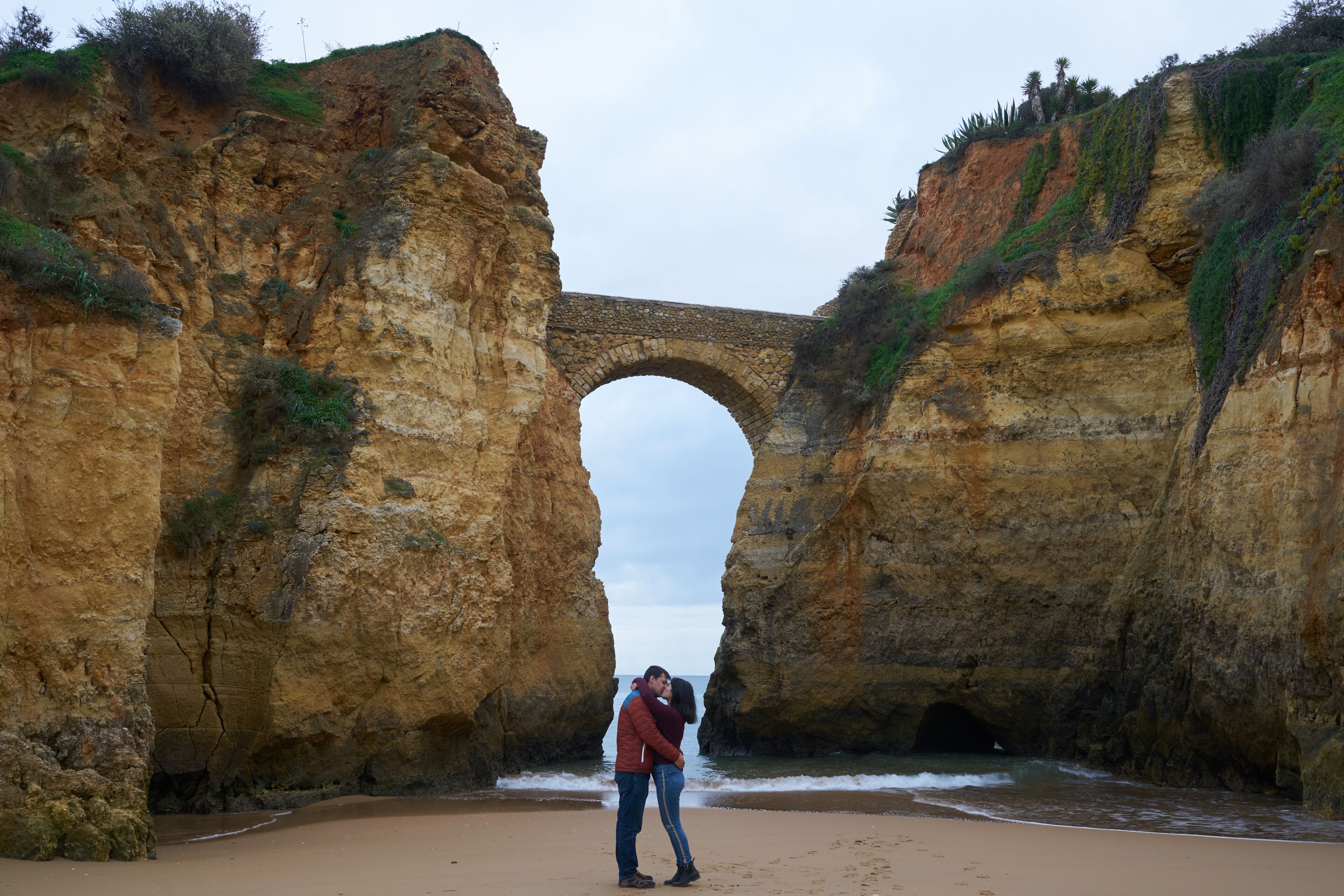 Um homem abraçando uma mulher em frente a um grande arco de pedra na praia.