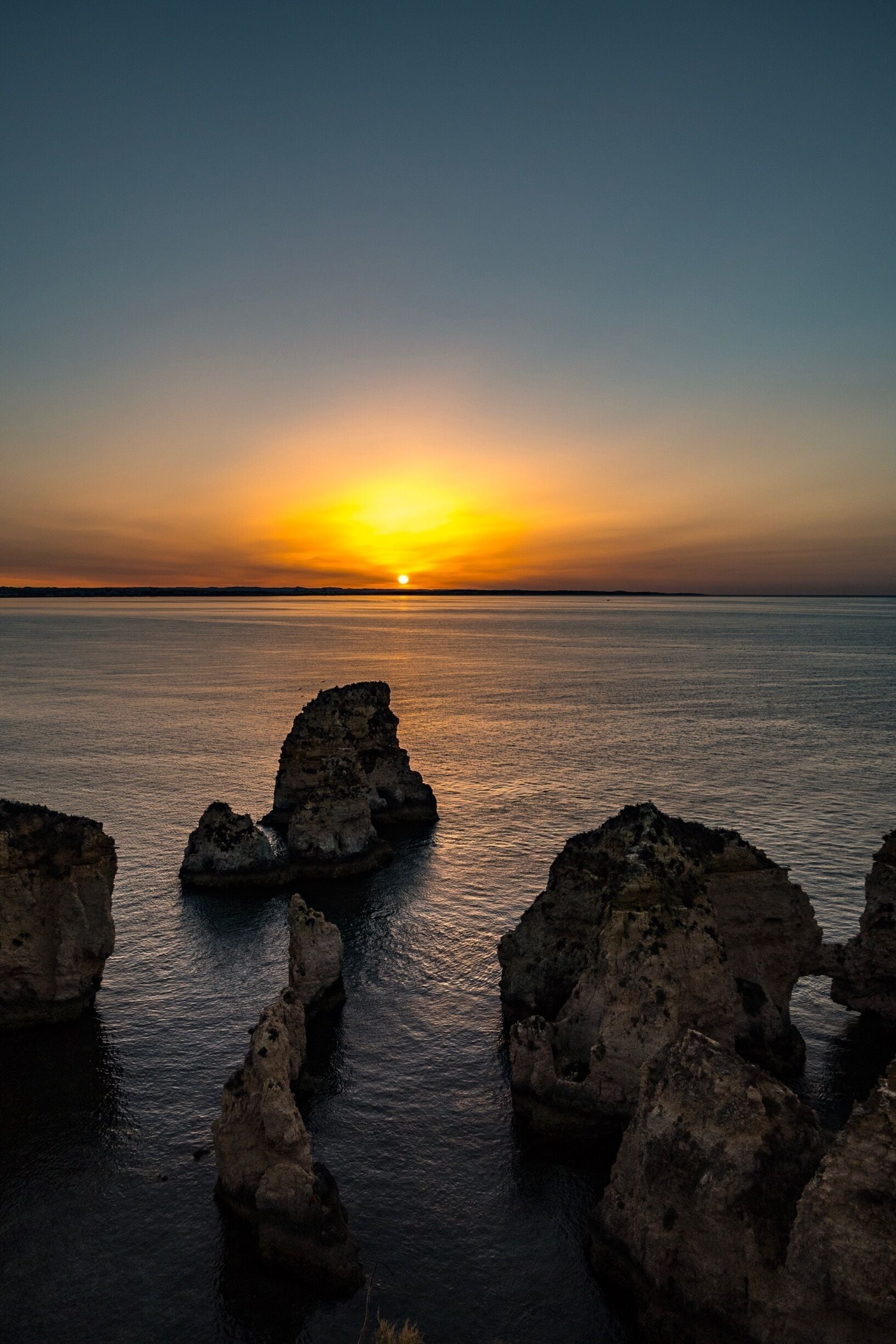 Um pôr do sol sobre um mar tranquilo, com formações rochosas em primeiro plano.