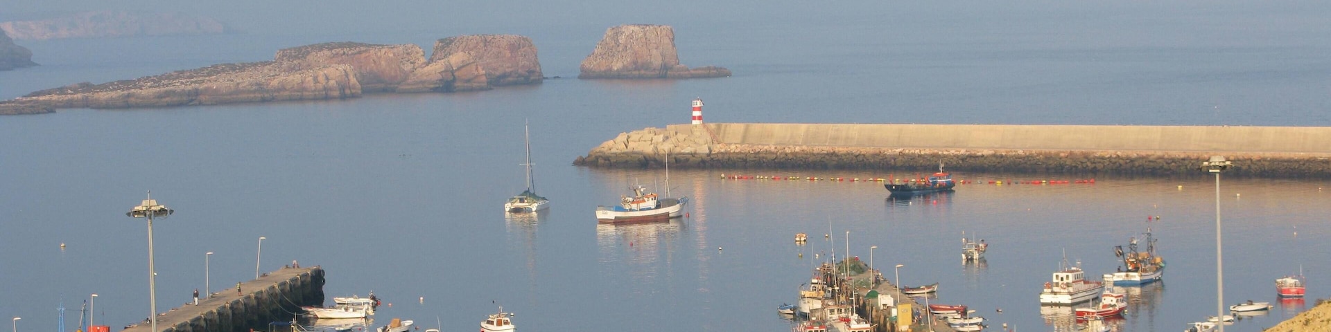 Um porto com muitos barcos atracados, um farol e formações rochosas ao fundo.