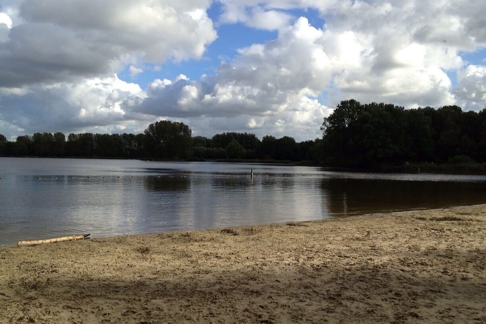 Um lago com margem arenosa, rodeado por árvores sob um céu parcialmente nublado.