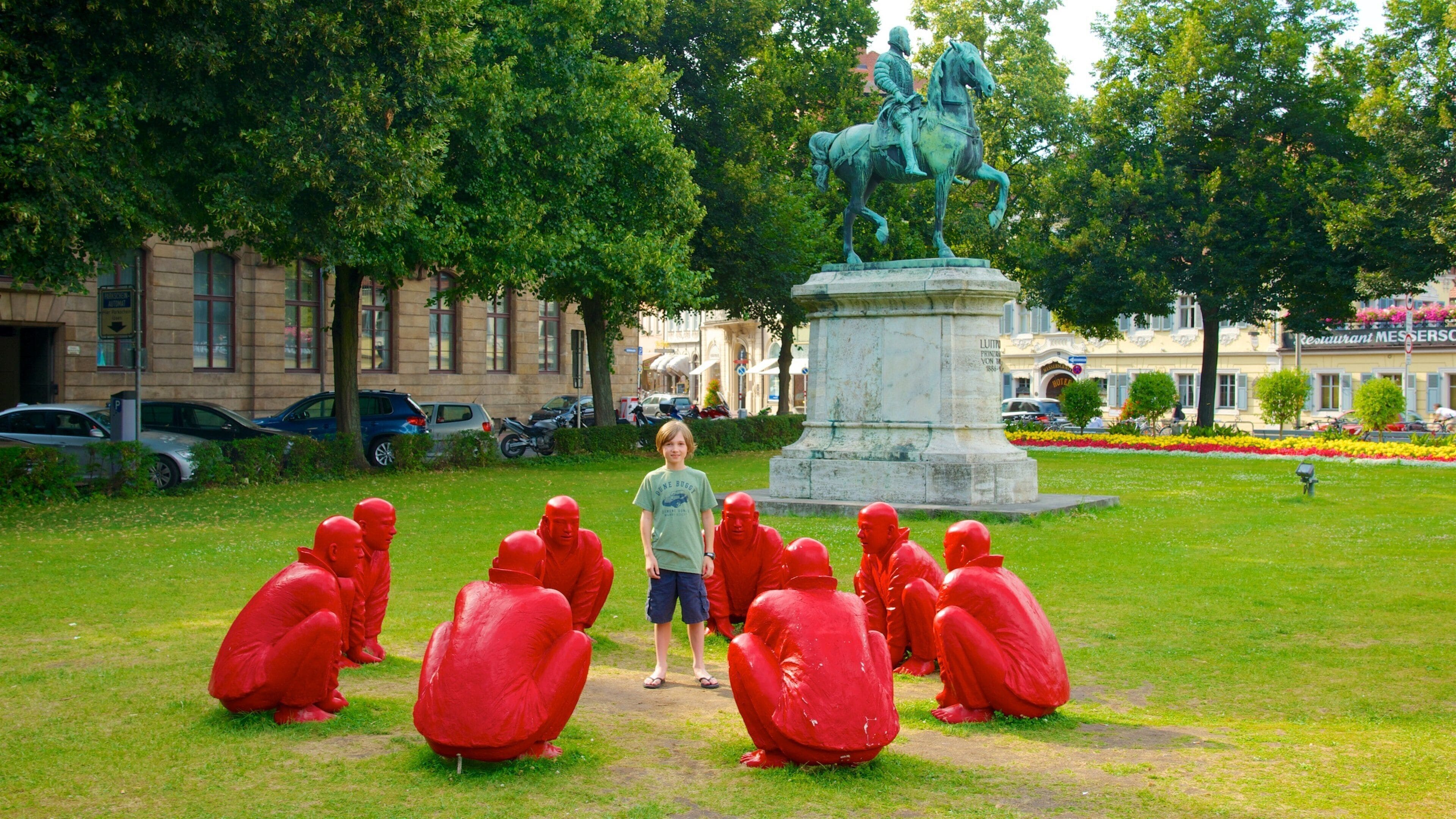 Uma estátua de um homem a cavalo, cercada por esculturas vermelhas, com um edifício e carros estacionados ao fundo.