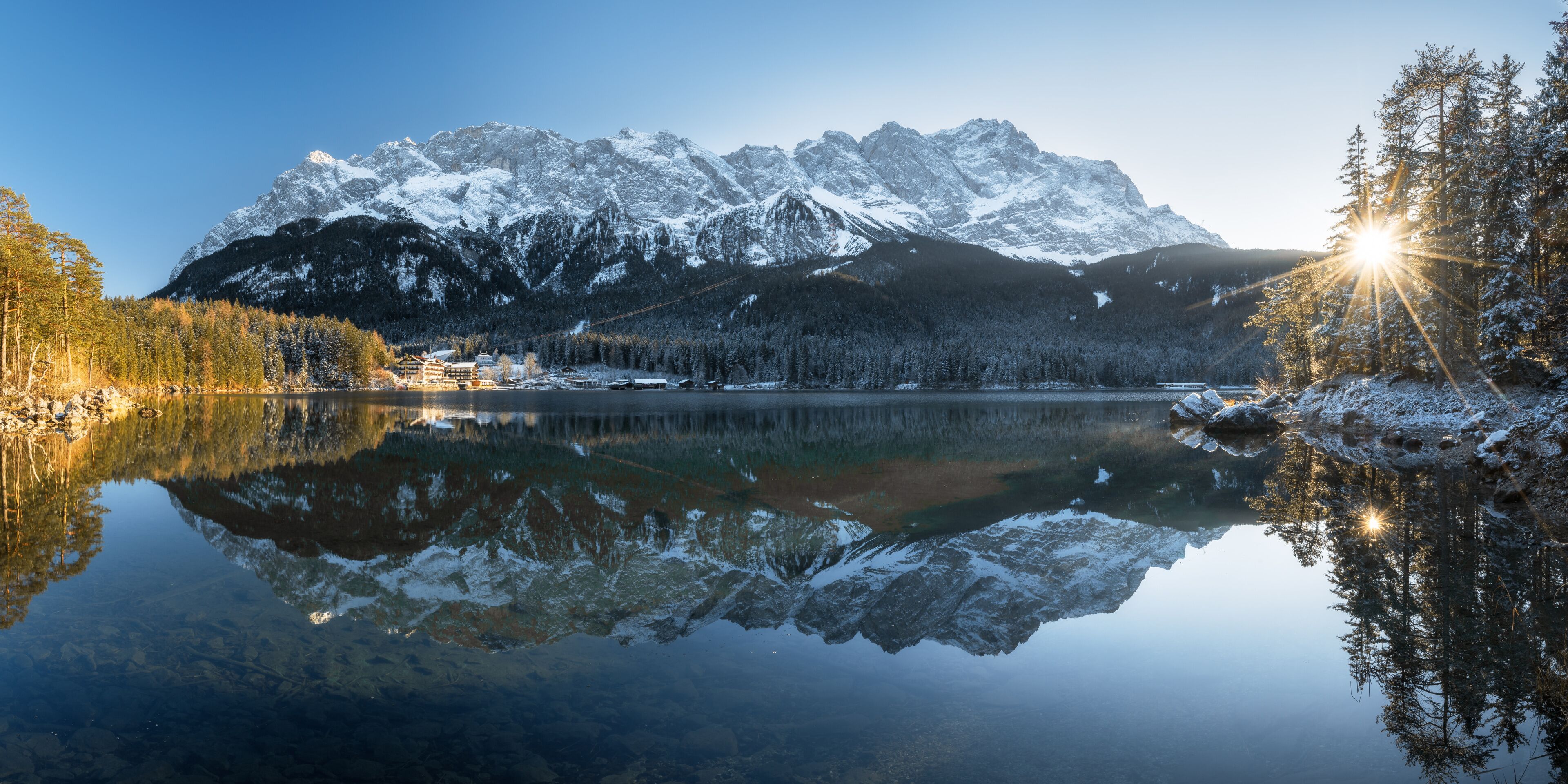 Cordilheira coberta de neve, um lago calmo refletindo a paisagem e uma área arborizada com edifícios.