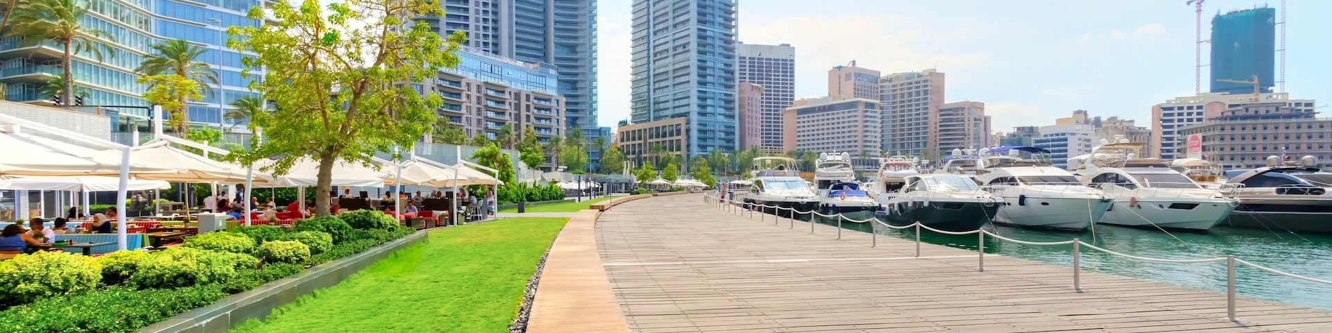 Um calçadão à beira-mar com edifícios modernos, uma área gramada e iates atracados.