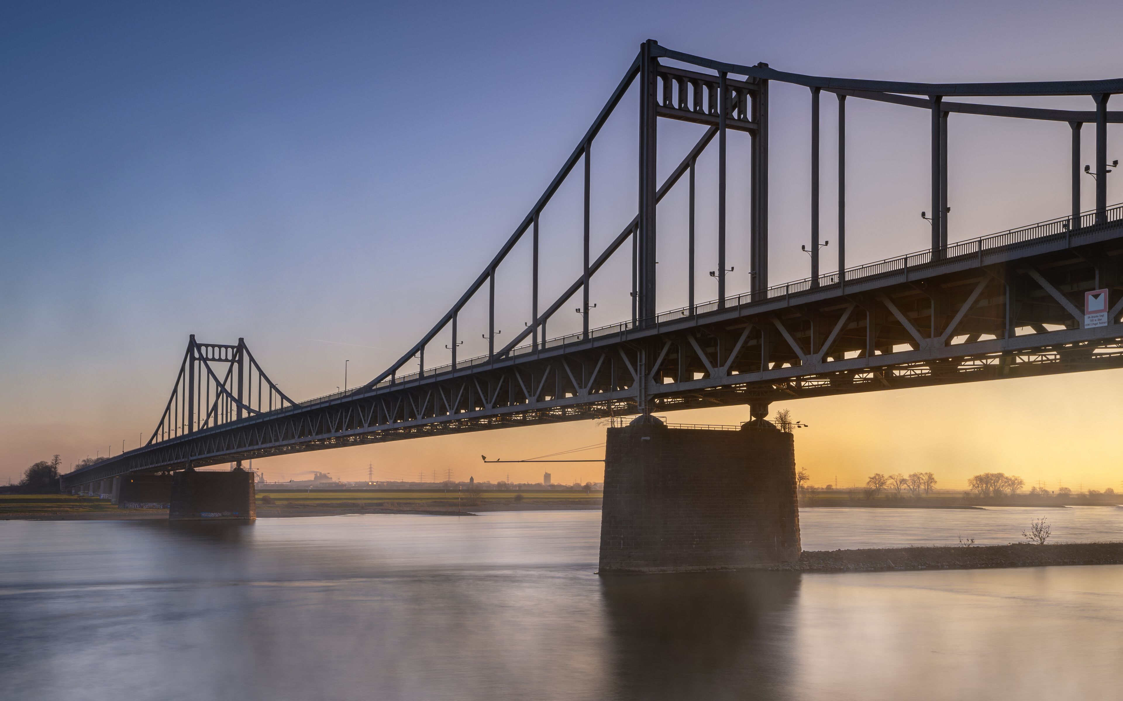 Uma grande ponte suspensa atravessa um rio amplo ao pôr do sol.
