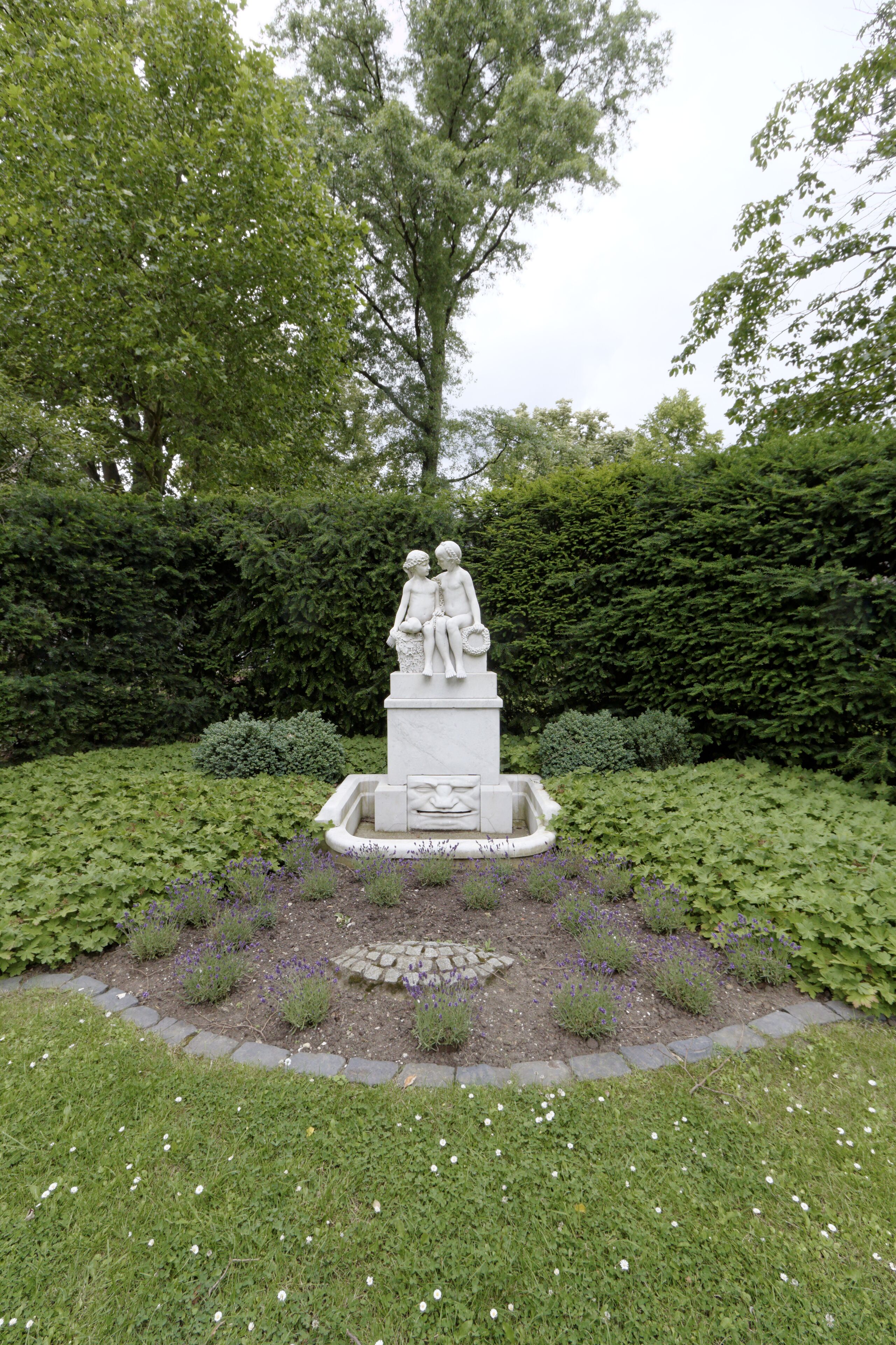 Estátua de duas figuras em um jardim com uma fonte, cercada por vegetação e plantas floridas.