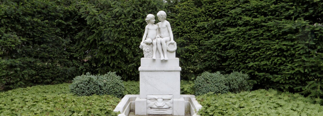 Estátua de duas figuras em um jardim com uma fonte, cercada por vegetação e plantas floridas.