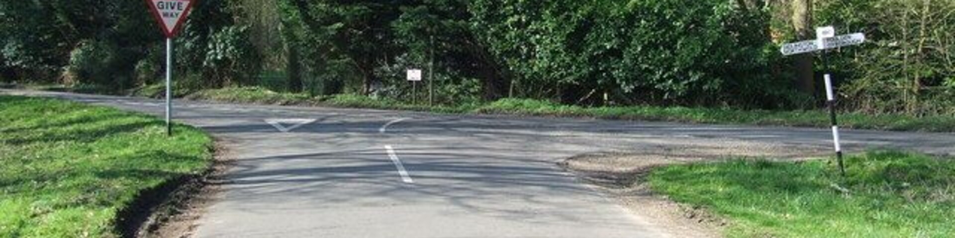 Cruzamento de estrada rural com placa de 'Dê a passagem' e placas de sinalização.