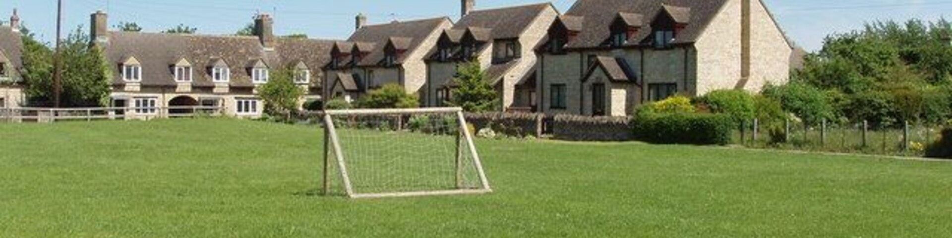 Um campo gramado com um gol de futebol, rodeado por casas com arquitetura tradicional.