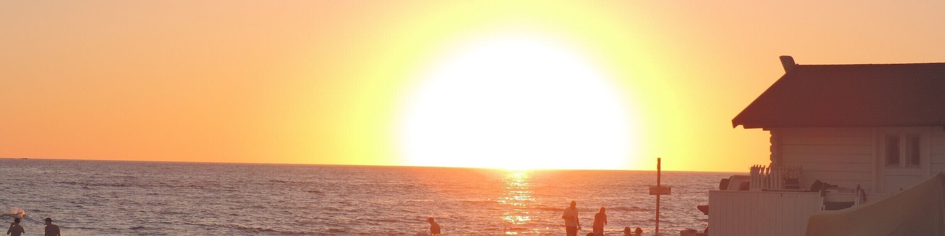 Um pôr do sol sobre o oceano, com pessoas na praia e um edifício em primeiro plano.