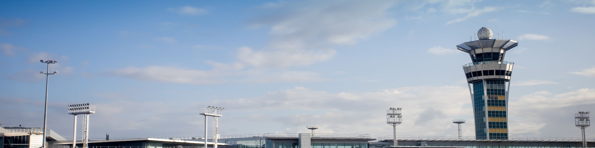 Um terminal de aeroporto com uma torre de controle, céu claro e pista com barreiras.
