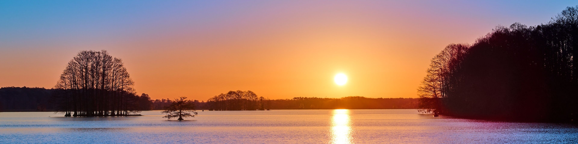 Um pôr do sol sobre um lago calmo, com árvores recortadas contra o céu.