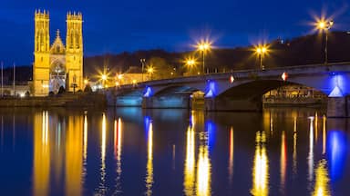 Uma ponte iluminada sobre um rio calmo que reflete luzes, com uma catedral histórica ao fundo.