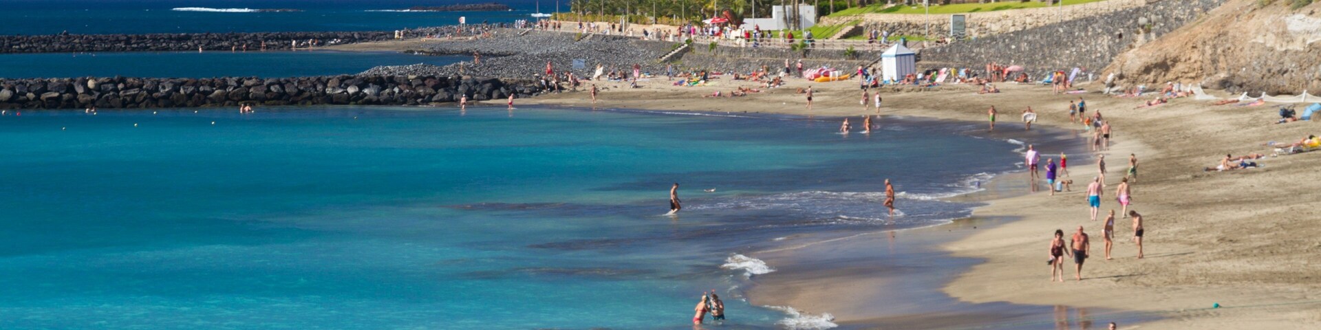 Uma praia movimentada com águas azul-turquesa cristalinas, palmeiras e um molhe rochoso.