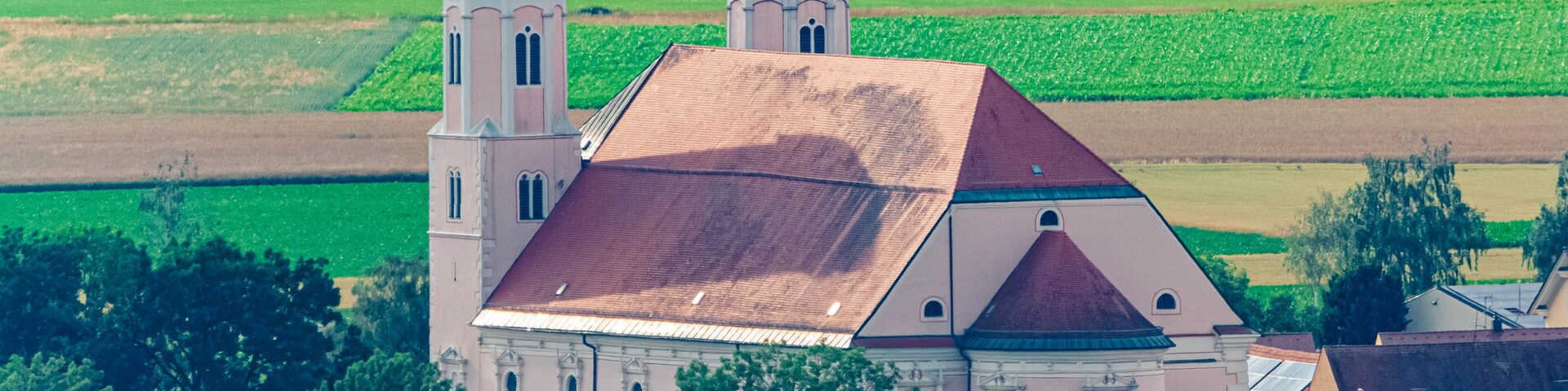 Igreja com duas torres e telhado vermelho, cercada por campos verdes e árvores.
