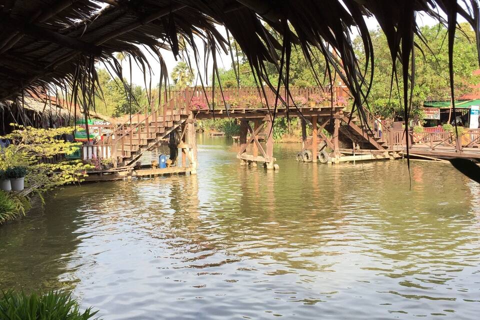Uma ponte de madeira sobre um rio calmo, cercada por vegetação exuberante e uma estrutura de telhado de palha.