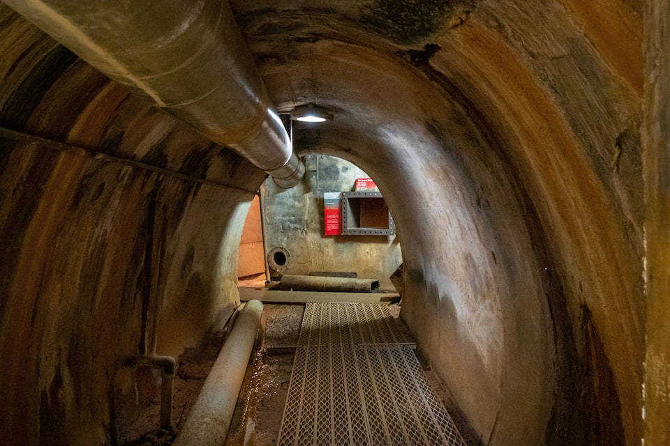 Um túnel subterrâneo com chão de grade metálica, canos e uma placa vermelha na parede.