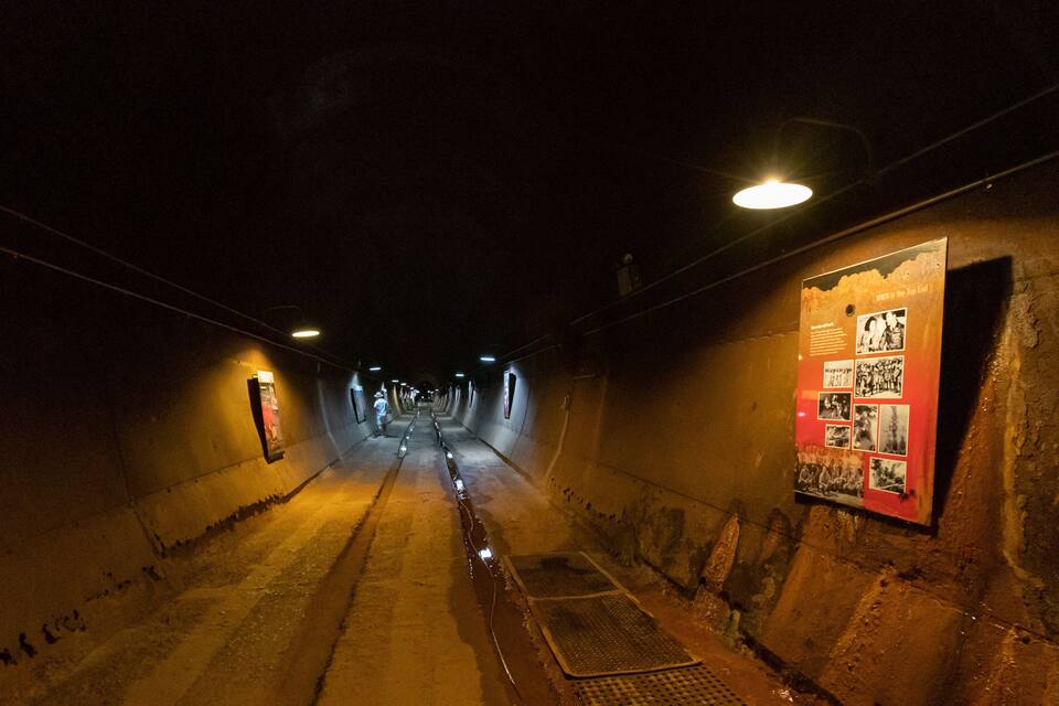 Um túnel pouco iluminado, com paredes de concreto, chão molhado e um cartaz na parede direita.