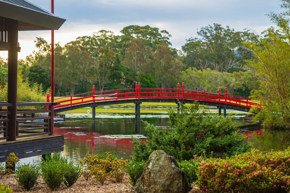 Uma ponte vermelha sobre um lago cercado por vegetação e um pavilhão de madeira.