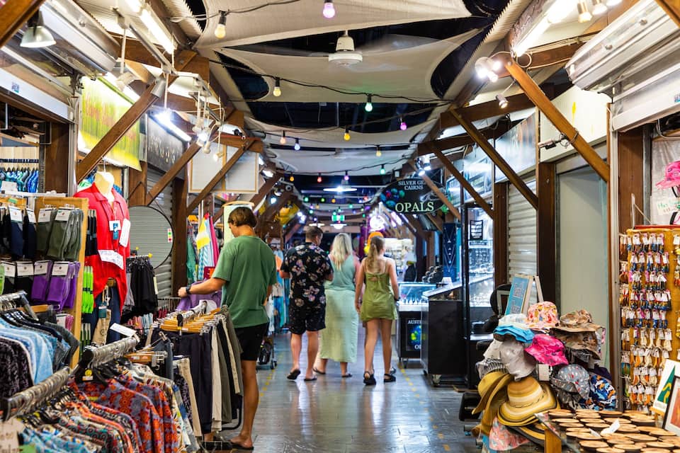 Uma passagem estreita em um mercado, com araras de roupas, chapéus e acessórios.