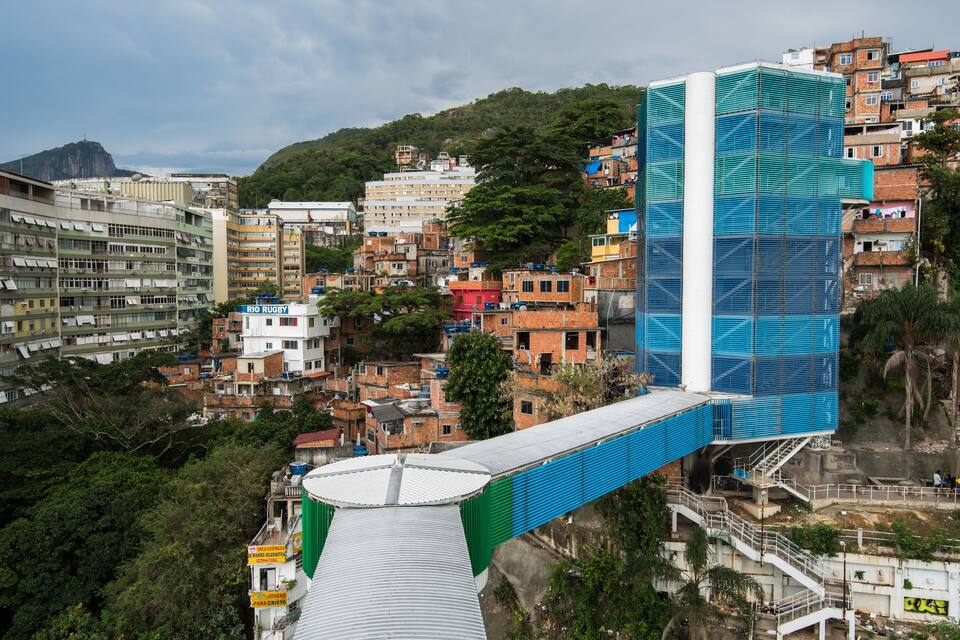 Paisagem urbana com um edifício azul e branco imponente, uma estrutura com telhado verde e montanhas ao fundo.