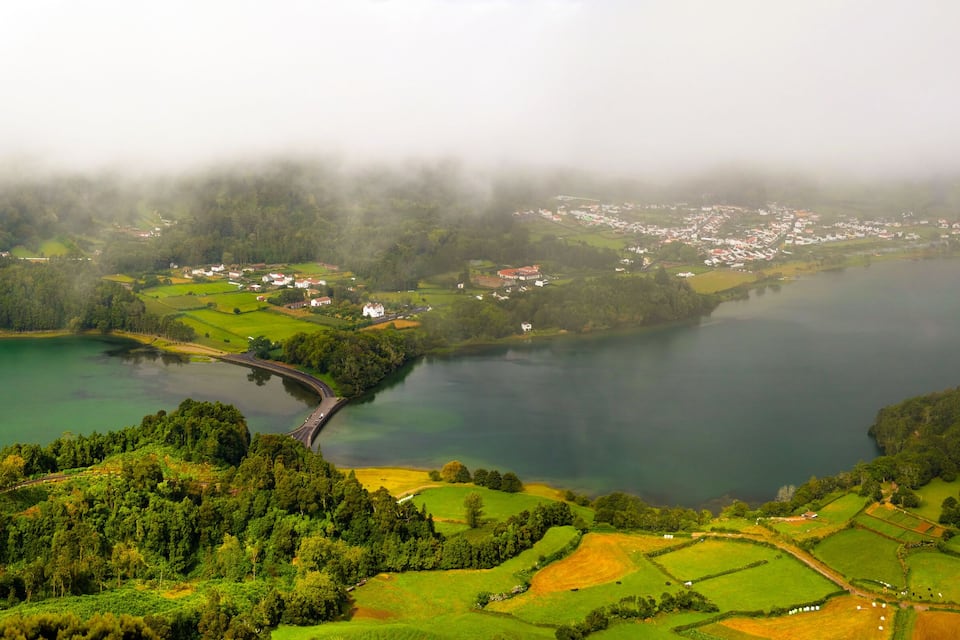 Paisagem nevoenta com um lago, campos verdes e uma pequena cidade.