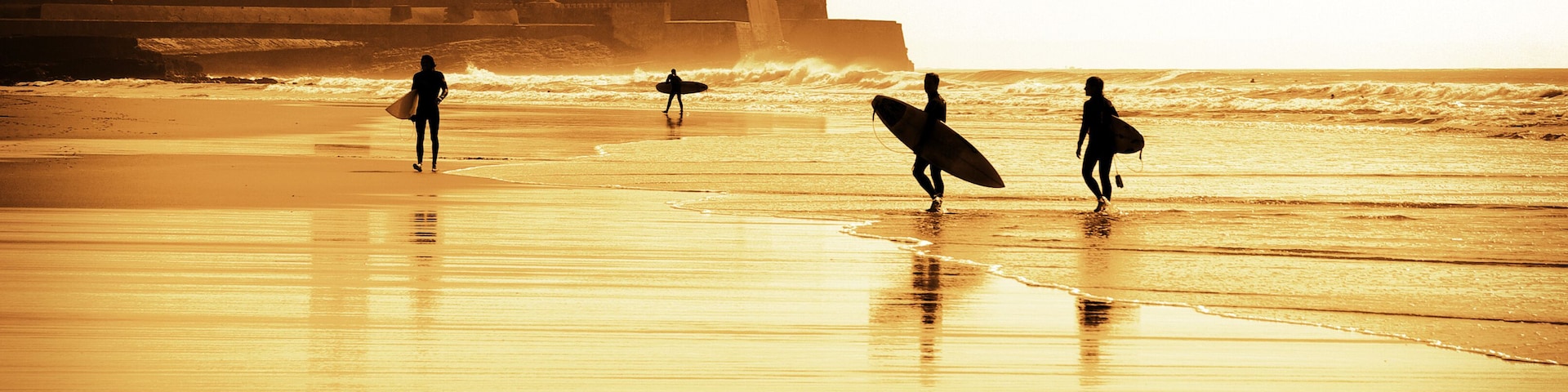 Uma praia com surfistas caminhando em direção à água, uma torre e ondas quebrando na costa.