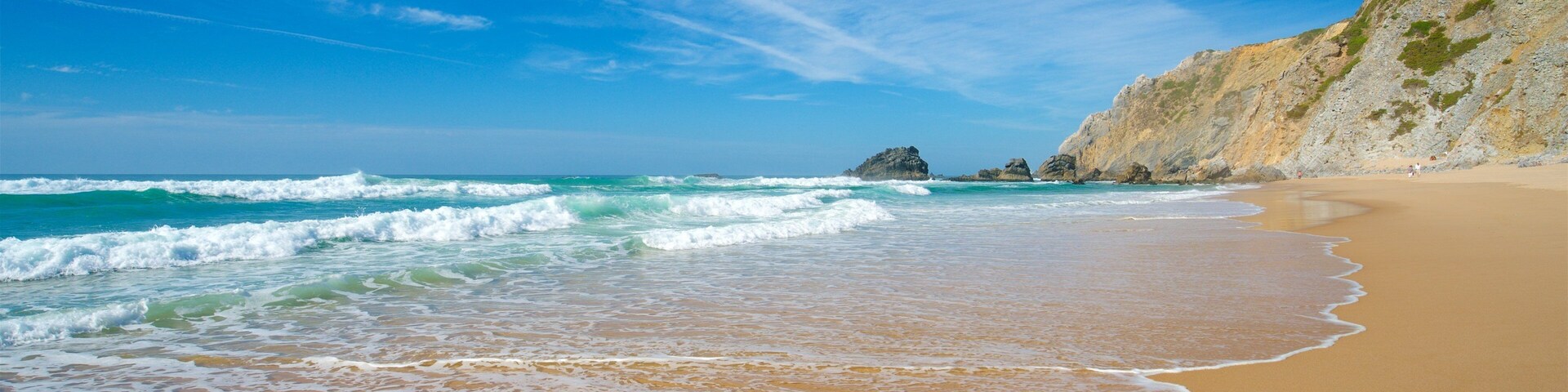 Uma praia arenosa com ondas, um penhasco rochoso e um céu azul límpido.