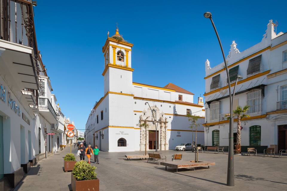 Uma praça com uma igreja branca e um campanário, bancos e uma rua com edifícios.