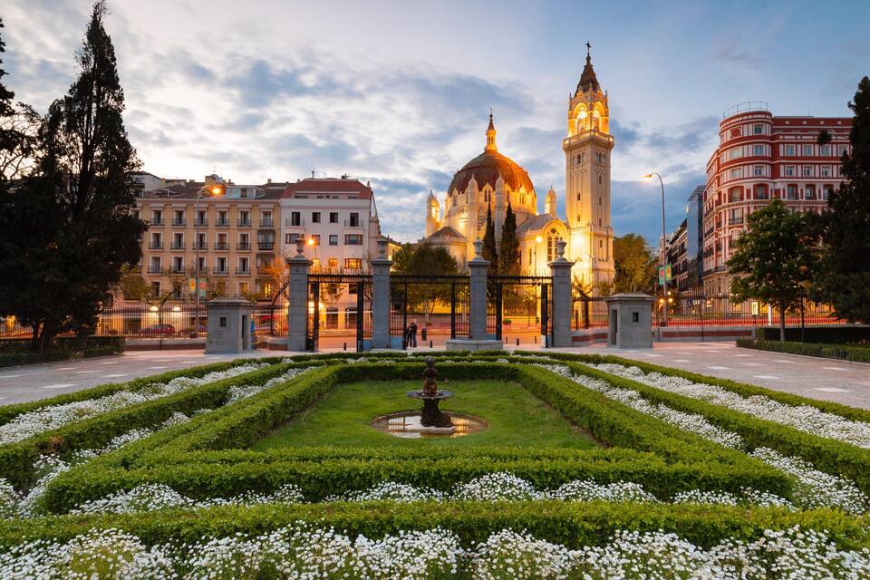 Uma praça urbana com uma fonte central, rodeada por jardins bem cuidados e uma catedral imponente com uma alta torre de sino.