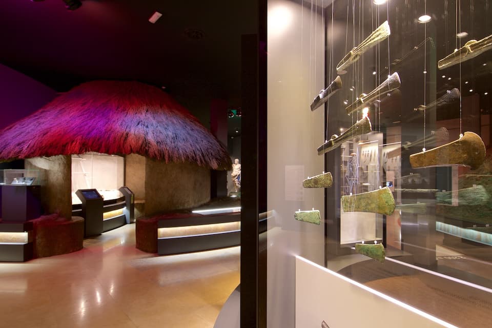Exposição de museu com uma cabana de colmo e diversos artefatos expostos em vitrines de vidro.