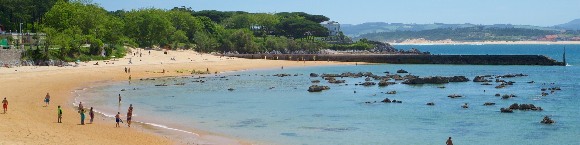 Uma praia de areia com águas claras, pedras e pessoas.