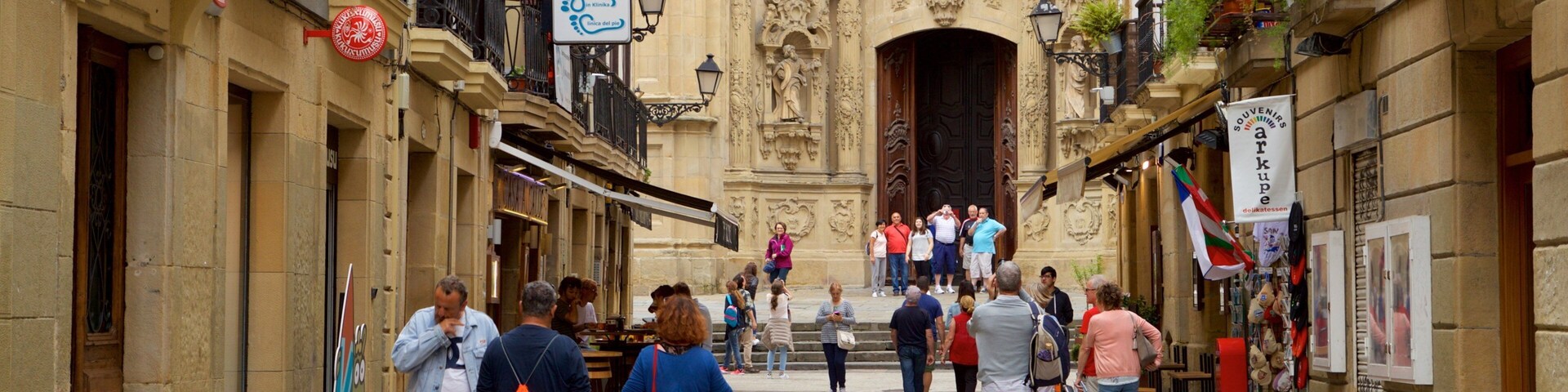 Rua estreita com edifícios ornamentados, uma entrada imponente e pessoas caminhando.