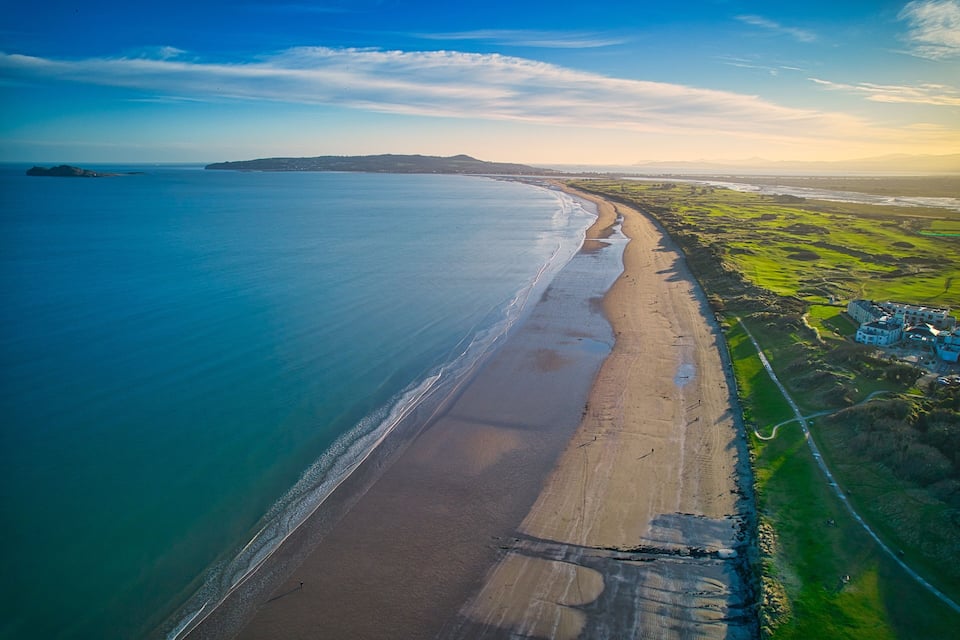 Paisagem costeira com uma praia arenosa, mar azul límpido e campos verdes.
