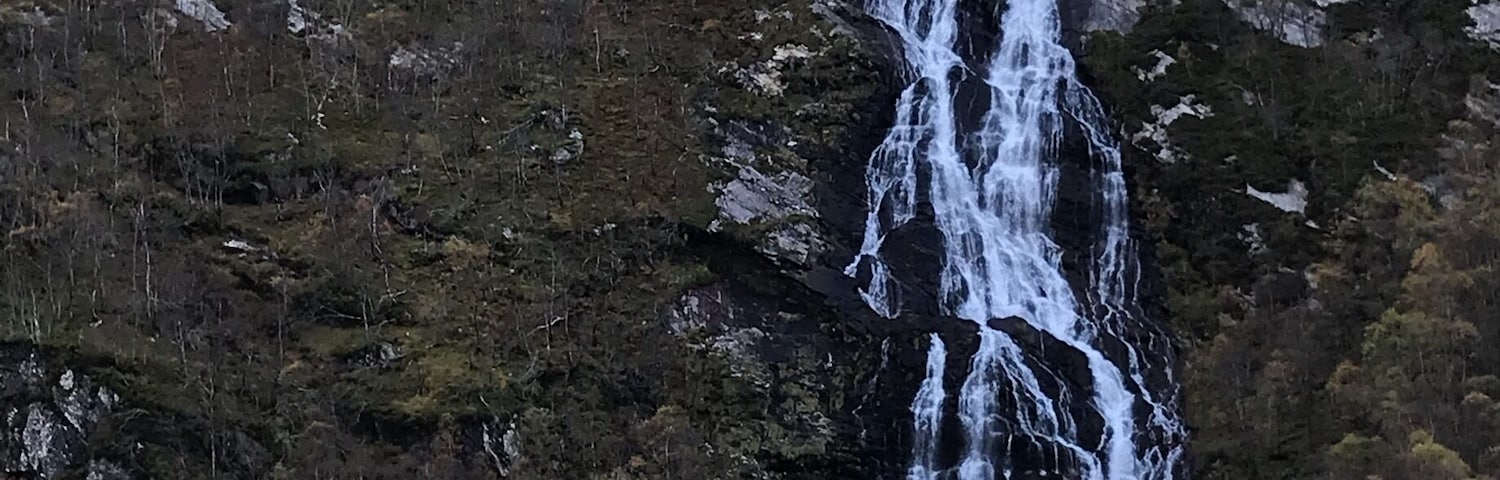 Uma cascata desce por uma montanha rochosa, com um campo em primeiro plano.