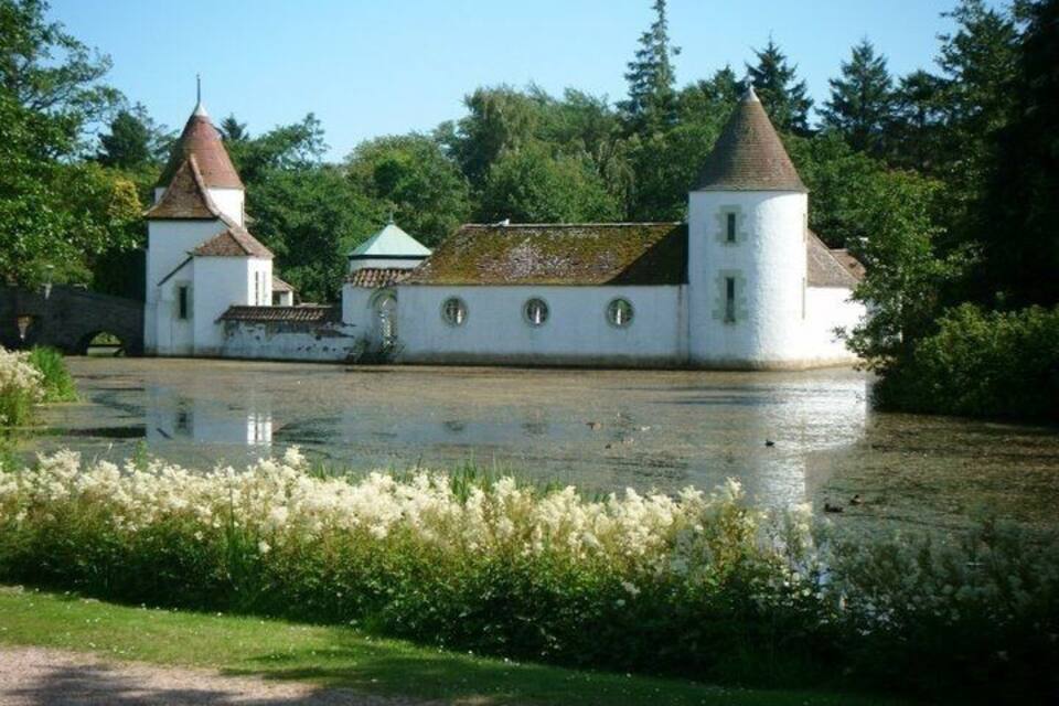 Uma igreja branca com um campanário e um edifício menor junto a um rio calmo.