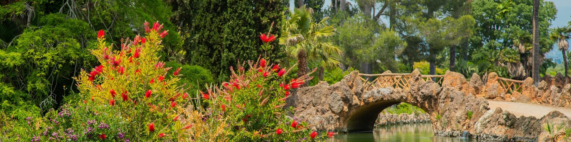 Uma ponte de pedra sobre um lago calmo, rodeada por vegetação exuberante e flores vibrantes.
