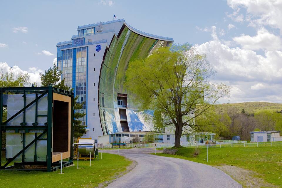 Um edifício moderno com fachada curva, rodeado por paisagem verde e céu claro.